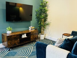 Sala de estar com unidade de entretenimento de madeira escura, TV de tela plana na parede, uma planta de bambu alta, sofá azul com almofadas estampadas e um tapete decorativo.