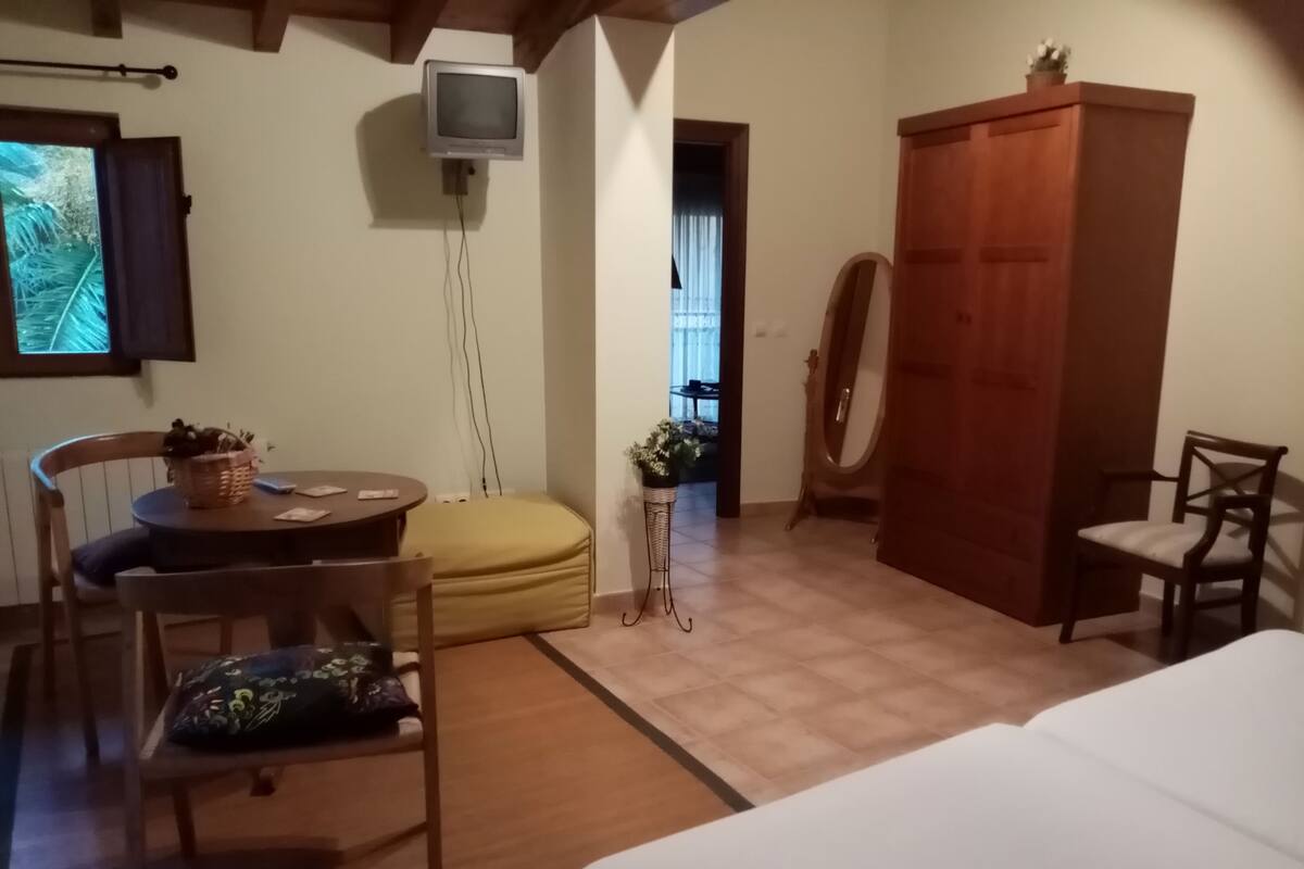 Quarto com cama, guarda-roupa de madeira, mesa pequena com cesta, cadeira e televisão fixada na parede.