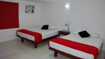 Quarto de hotel com duas camas, cada uma com colcha vermelha e lençóis brancos, mesa de cabeceira com abajur, relógio de parede e uma pequena planta em vaso.