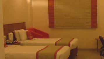 Quarto de hotel com três camas, um ventilador de teto, uma janela com persianas e uma mesa de cabeceira com abajur.