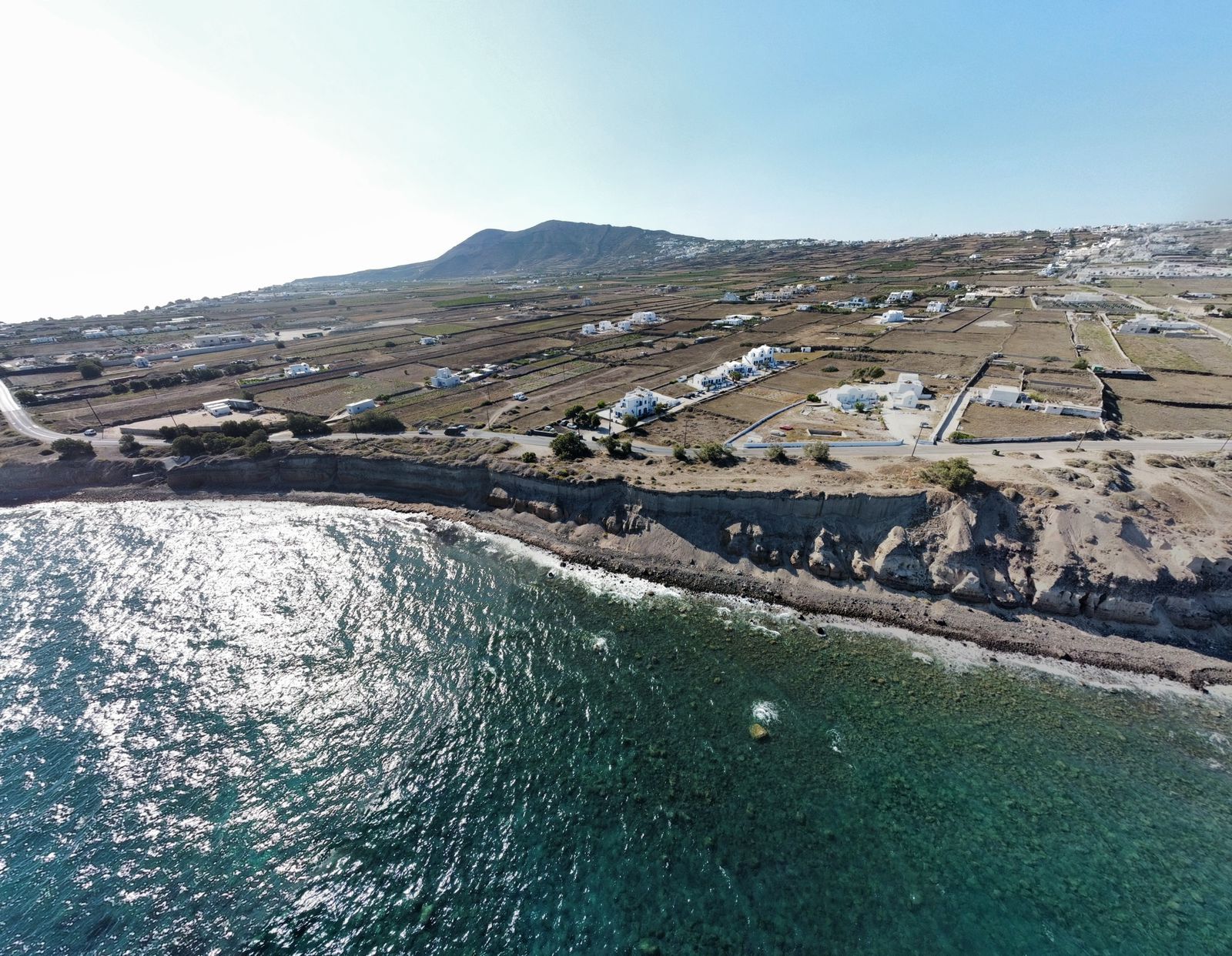 Vista aérea de uma área costeira com edifícios brancos, águas azul-claras e uma montanha ao longe.