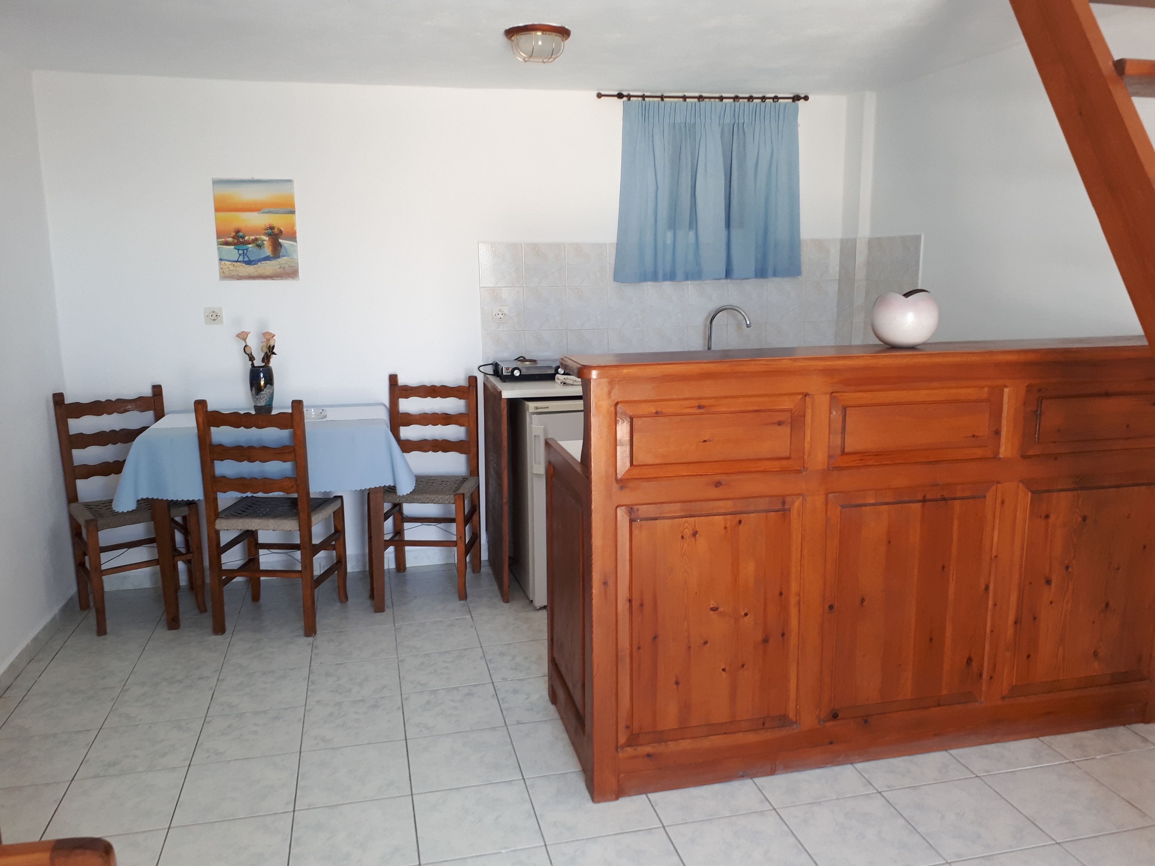 Cozinha compacta com ilha de madeira, área de jantar com duas cadeiras, mesa pequena e luminária fixada na parede.