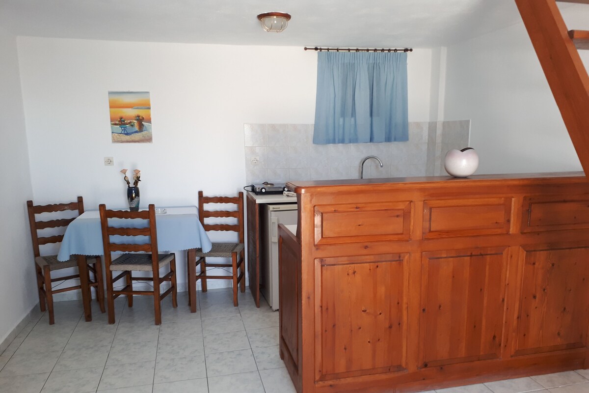 Cozinha compacta com ilha de madeira, área de jantar com duas cadeiras, mesa pequena e luminária fixada na parede.