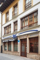 Fachada de hotel com várias janelas, uma varanda com grades de metal ornamentadas e uma placa indicando que é um hotel.