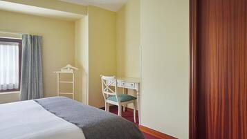 Quarto de hotel com cama, escrivaninha, cadeira, guarda-roupa e janela com cortinas.