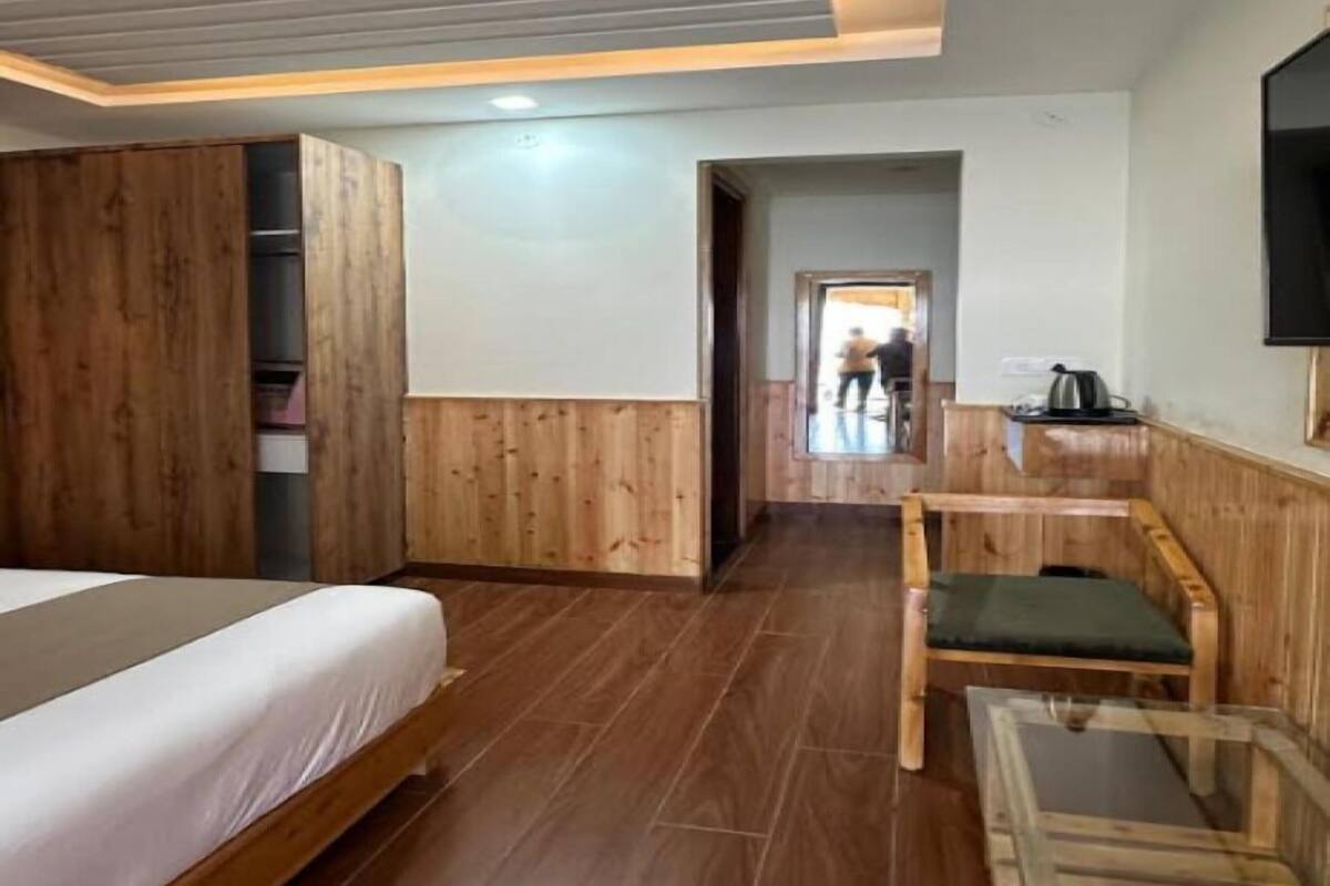 Quarto de hotel com cama, guarda-roupa de madeira, um espaço de estar pequeno e uma televisão na parede.