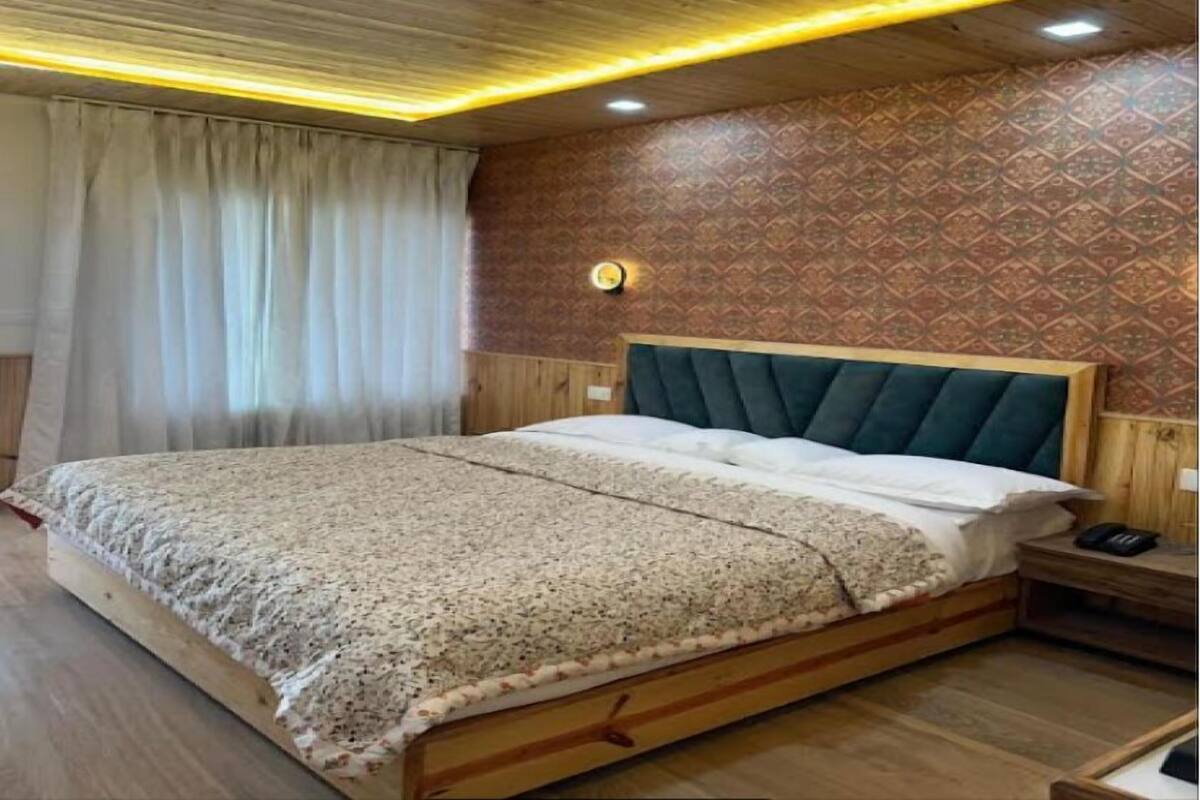 Quarto com teto de madeira, papel de parede estampado, cama com cabeceira verde, mesa de cabeceira e apoio para luminária.