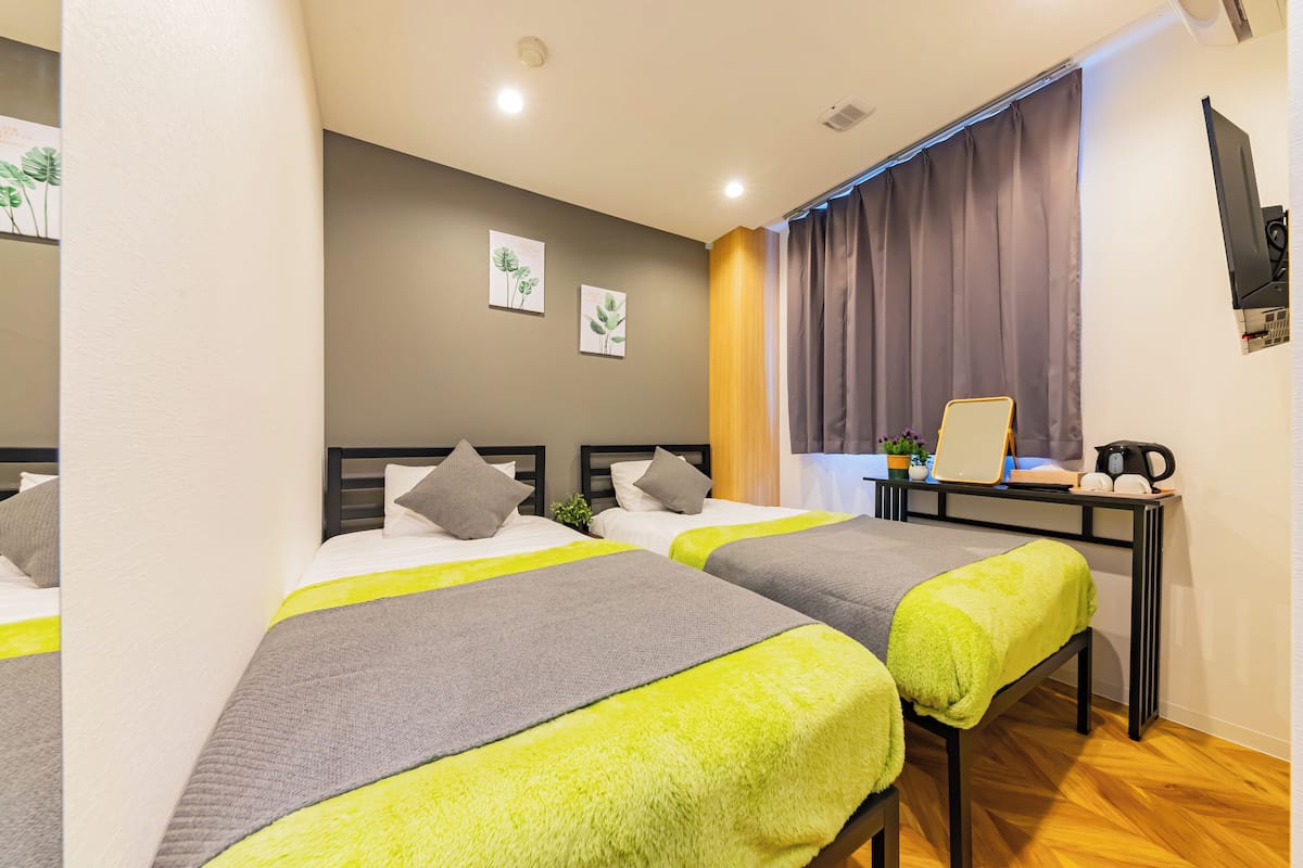 Quarto de hotel com duas camas, uma escrivaninha com espelho, cafeteira e janela com cortinas.