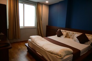 Quarto de hotel com uma cama grande, cabeceira de madeira, janela com cortinas e piso de madeira.