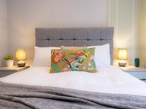 Cama bem arrumada com cabeceira estofada cinza, duas luminárias de apoio e um travesseiro decorativo com motivos de pássaros e flores.