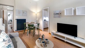 Uma sala de estar moderna com televisão de tela plana, área de jantar com cadeiras, mesa de centro com xícaras e pires, e cozinha ao fundo.