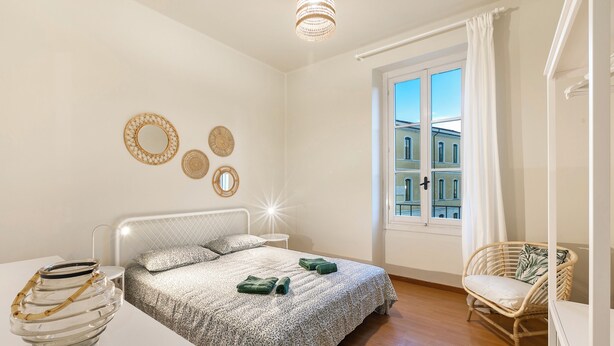 Quarto com cama, mesa de cabeceira, arte decorativa na parede, janela com cortinas e uma cadeira de vime.