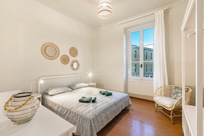 Quarto com cama, mesa de cabeceira, espelhos decorativos na parede, uma cadeira de vime e uma janela com vista para prédios.