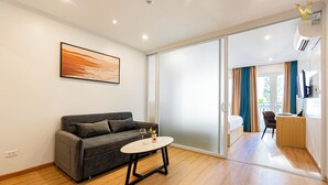 Uma sala de estar moderna com um sofá cinzento, uma mesa redonda pequena e um painel de vidro que dá para uma varanda com uma cadeira e uma escrivaninha.