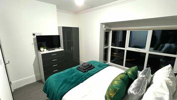 Um quarto com cama, televisão sobre cômoda, guarda-roupa e janela.