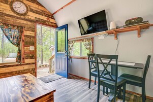 Interior rústico de cabana com teto de madeira, porta azul, televisão presa a uma prateleira de madeira e mesa de jantar com cadeiras.