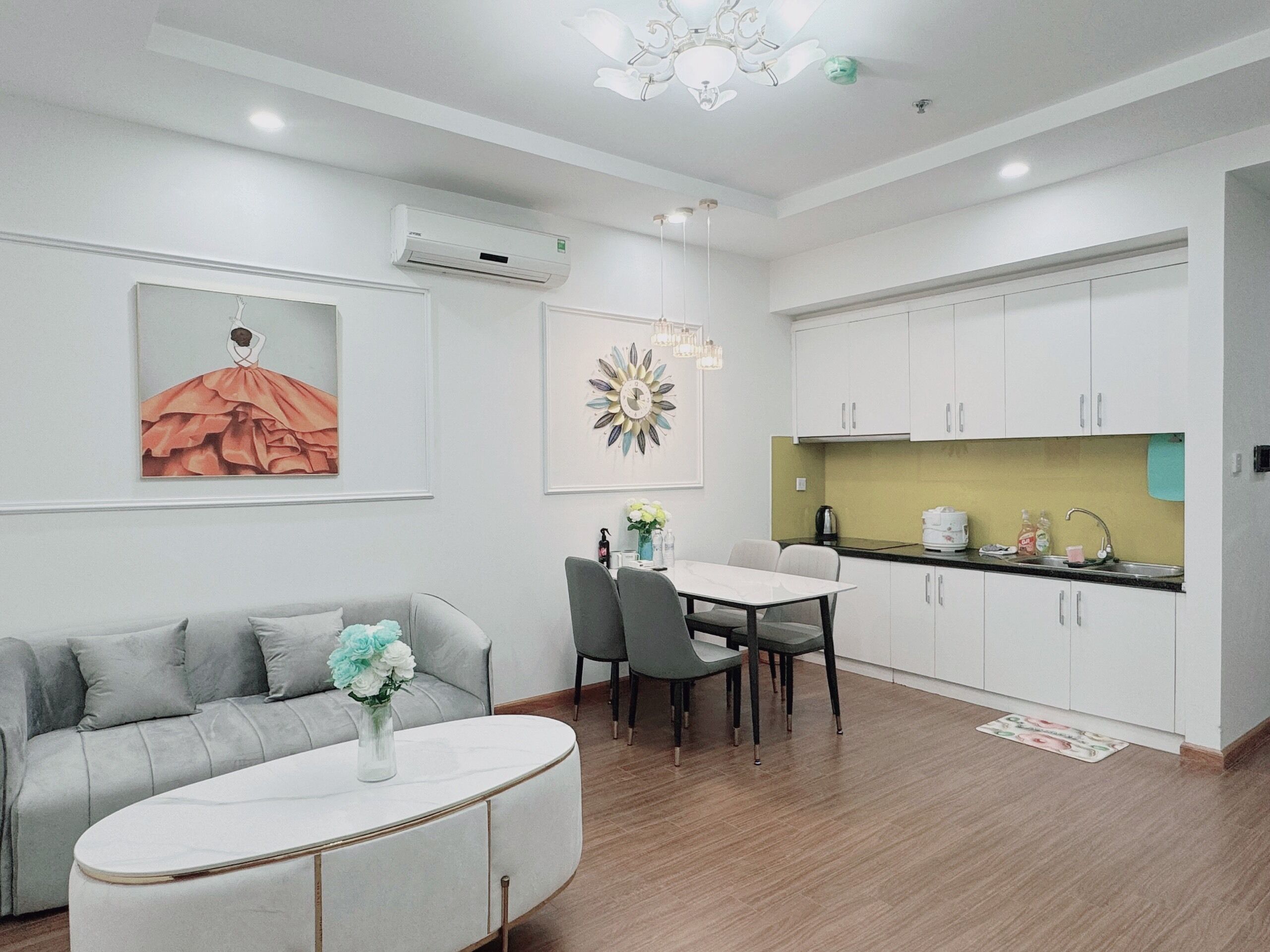 Uma sala de estar moderna com um sofá cinza, uma mesa de centro redonda, uma área de jantar com mesa branca e cadeiras cinzas, e uma cozinha com armários brancos e painel de fundo amarelo.
