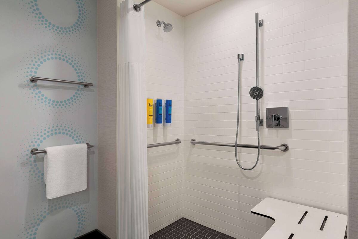 Casa de banho moderna com paredes revestidas de azulejos brancos, chuveiro com chuveiro manual e barra de apoio. Inclui um porta-toalhas com toalha branca e duas embalagens de shampoo.
