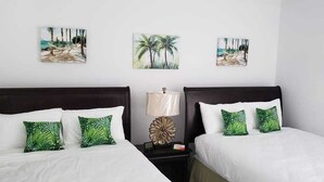 Quarto de hotel com duas camas, roupa de cama branca, almofadas estampadas em verde, mesa de cabeceira com abajur e três obras de arte nas paredes retratando paisagens tropicais.