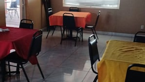 Uma área de refeições com mesas cobertas por toalhas vermelhas e amarelas, cadeiras pretas e uma janela com persianas.