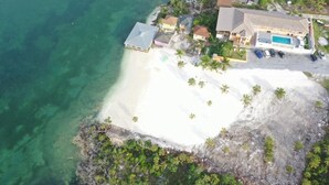 Vista aérea de uma propriedade costeira com uma extensa praia de areia, uma piscina e vários edifícios.