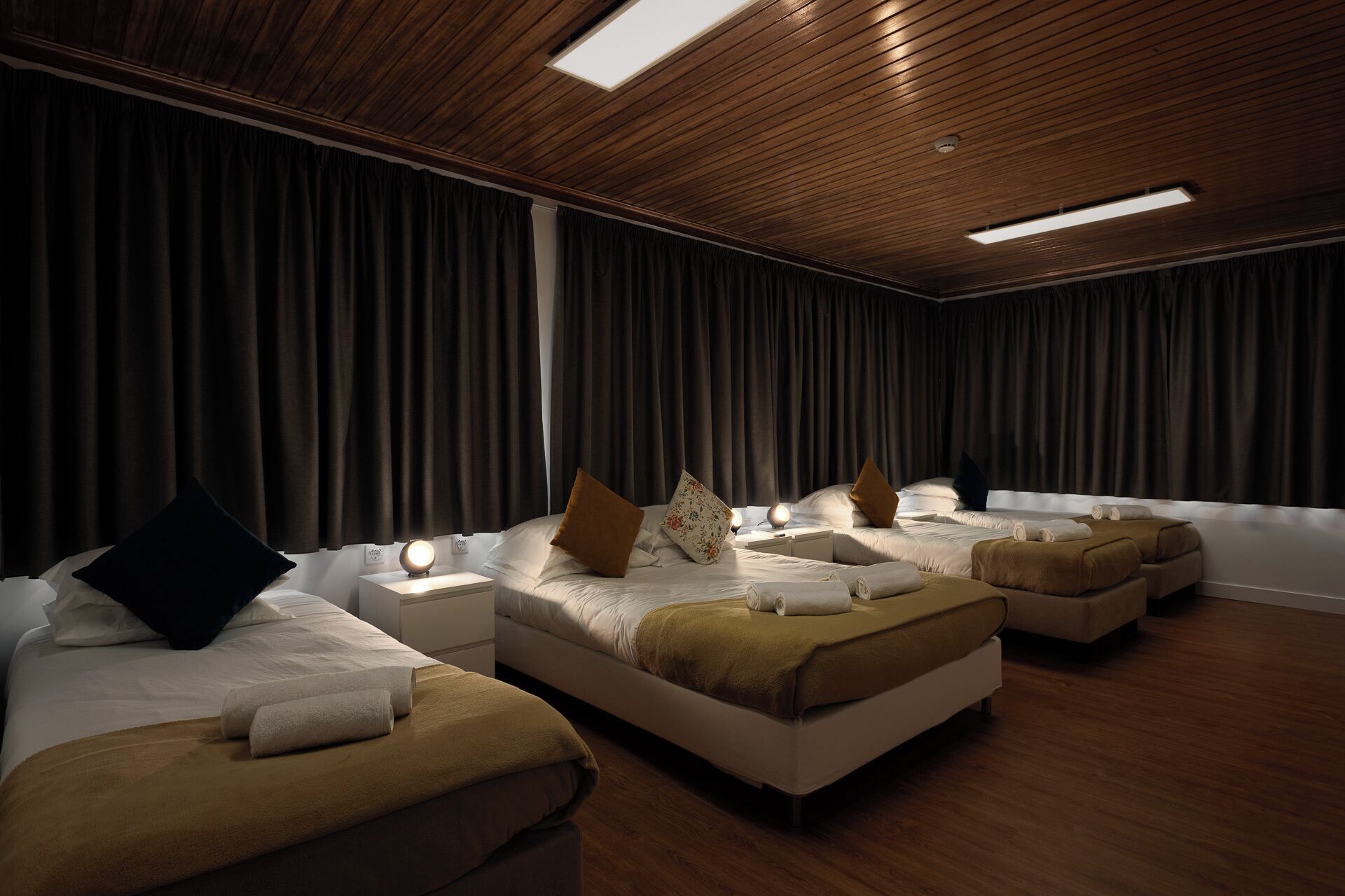 Quarto de hotel com teto de madeira, janelas amplas com cortinas escuras e várias camas com toalhas e almofadas.
