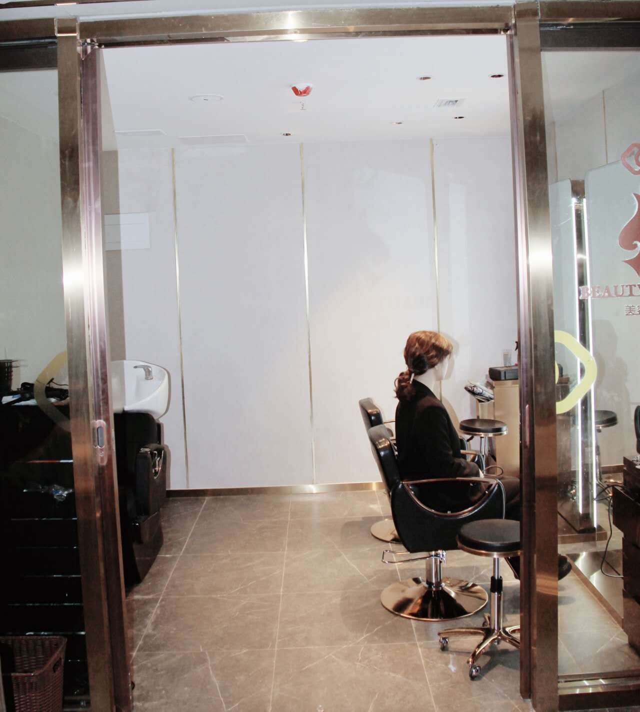 Interior de salão de beleza com uma única cliente sentada em uma cadeira preta, uma cuba branca e um espelho com um logotipo.