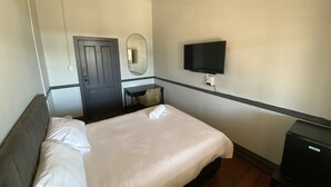 Quarto de hotel com cama, secretária, cadeira, televisão e porta.