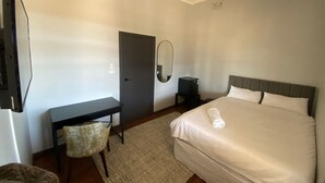 Quarto de hotel com cama, escrivaninha, cadeira, espelho e televisão.