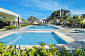 Uma piscina retangular com água azul cristalina, rodeada por vegetação e edifícios modernos.