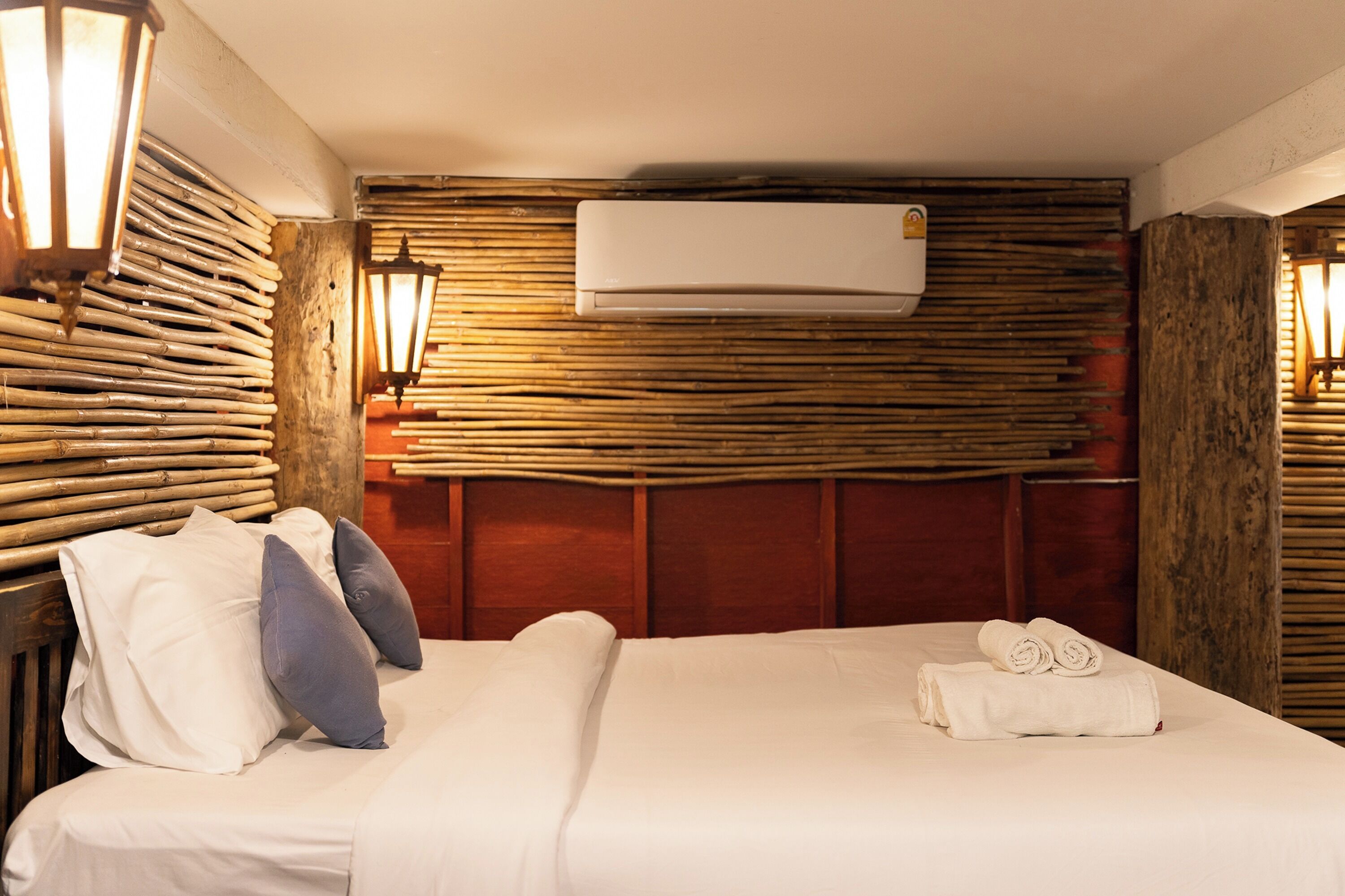 Quarto de hotel com cama, travesseiros, ar condicionado fixado na parede e duas arandelas.