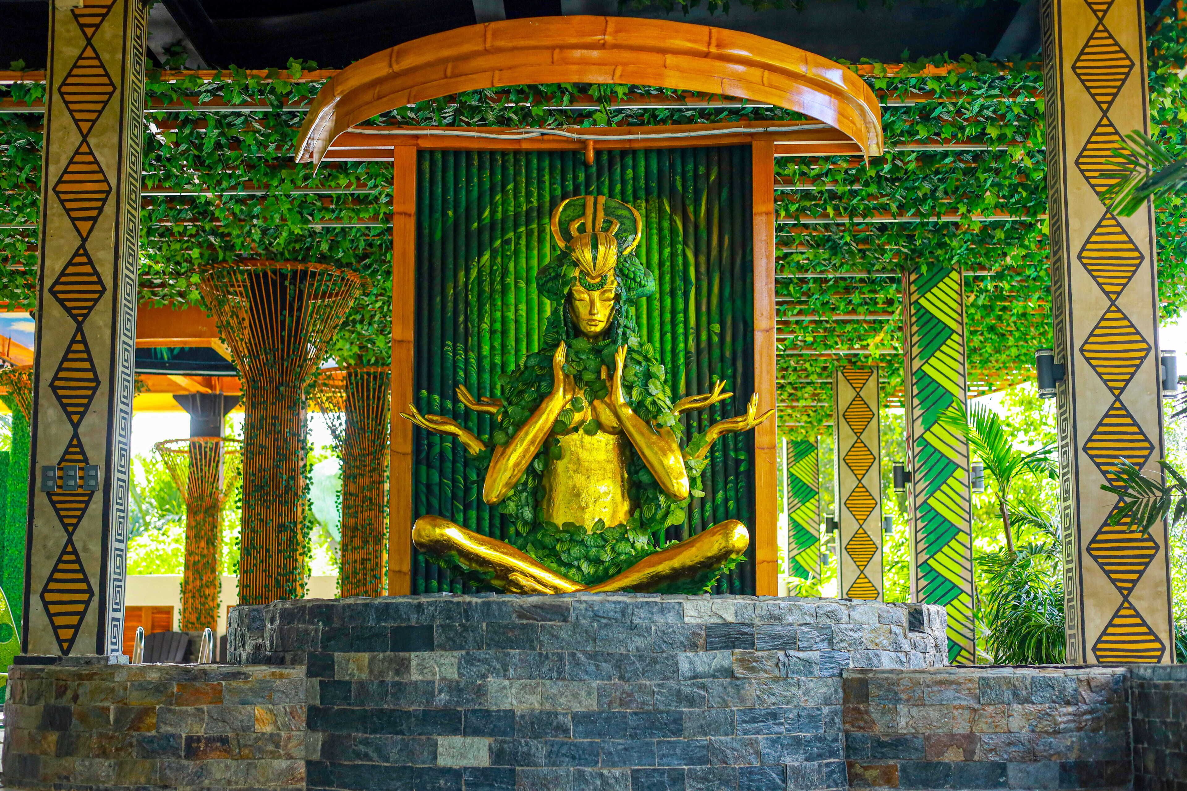 Uma estátua de uma figura em posição meditativa, cercada por vegetação e padrões tradicionais.
