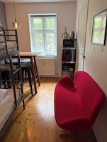 Quarto pequeno com beliches, uma cadeira vermelha, janela com vista, radiador e micro-ondas.