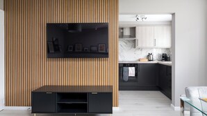 Uma sala de estar moderna com um móvel de entretenimento preto, uma parede de destaque em madeira e uma cozinha ao fundo.
