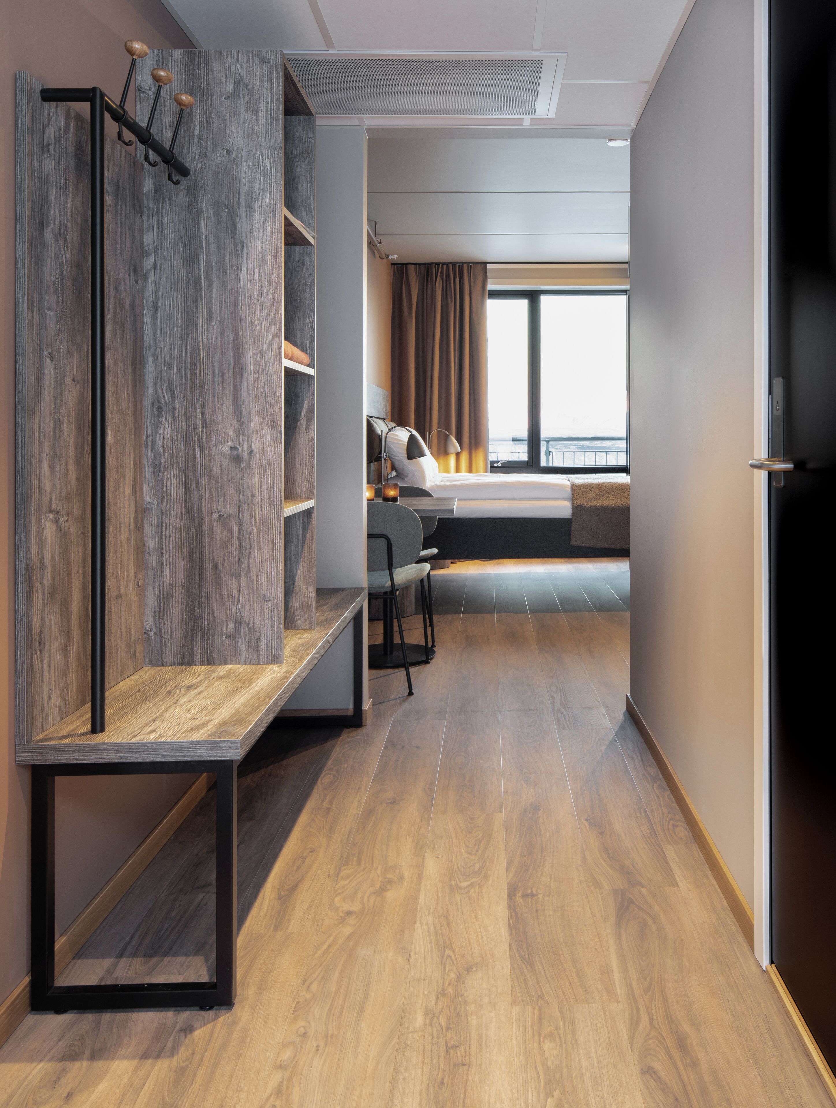 Quarto de hotel moderno com piso de madeira, uma escrivaninha com cadeira, uma cama e uma janela com cortinas.