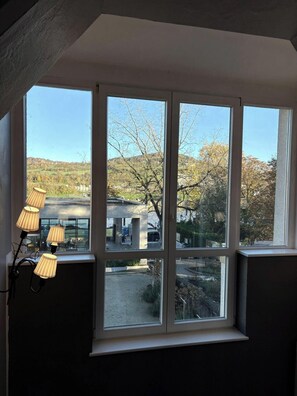 Quarto com uma janela ampla que dá para um parque e um edifício. Há três luminárias em um suporte à esquerda da janela.