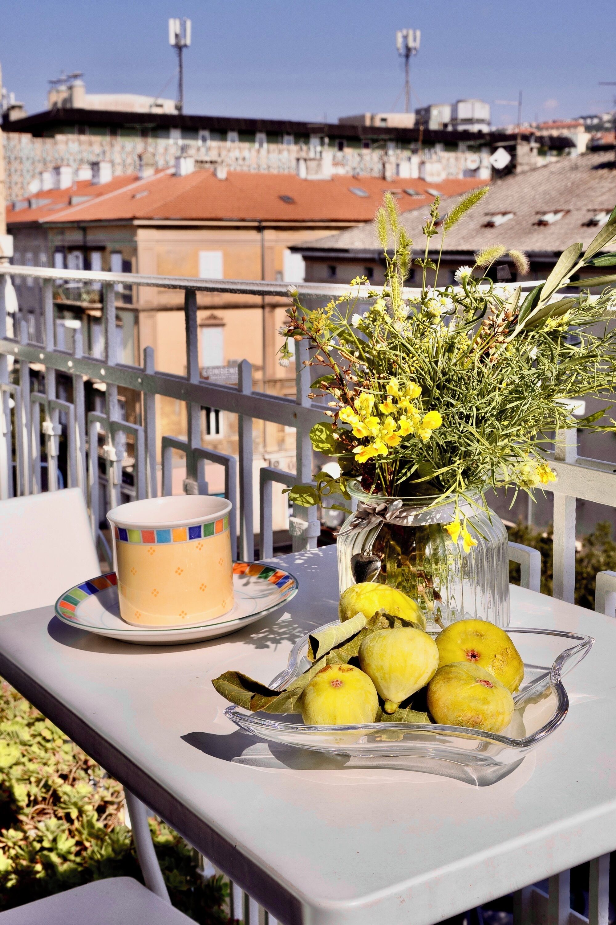 Uma mesa com um vaso de flores amarelas, um prato com limões e uma xícara em uma varanda com vista para edifícios.