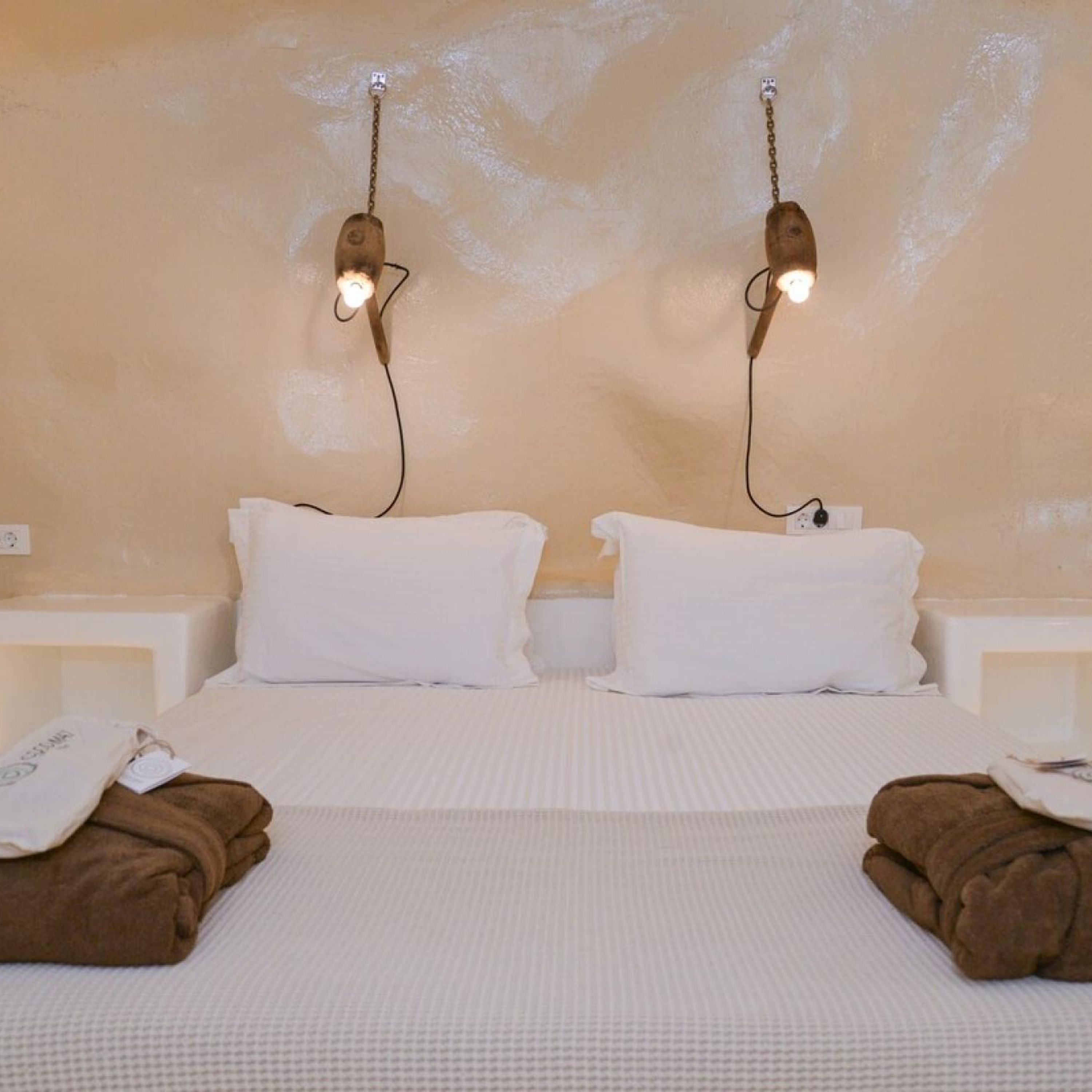 Quarto de hotel com duas camas, roupa de cama branca e duas luminárias na parede.