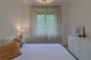 Um quarto com uma cama, mesa de cabeceira, luminária, janela com cortinas leves e um armário com moldura de quadro.