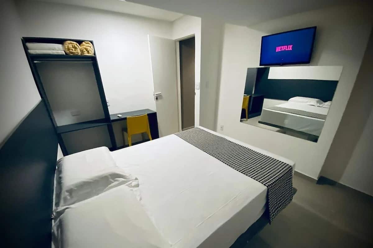 Quarto de hotel com cama, televisão fixada na parede, pequena escrivaninha e uma cadeira.