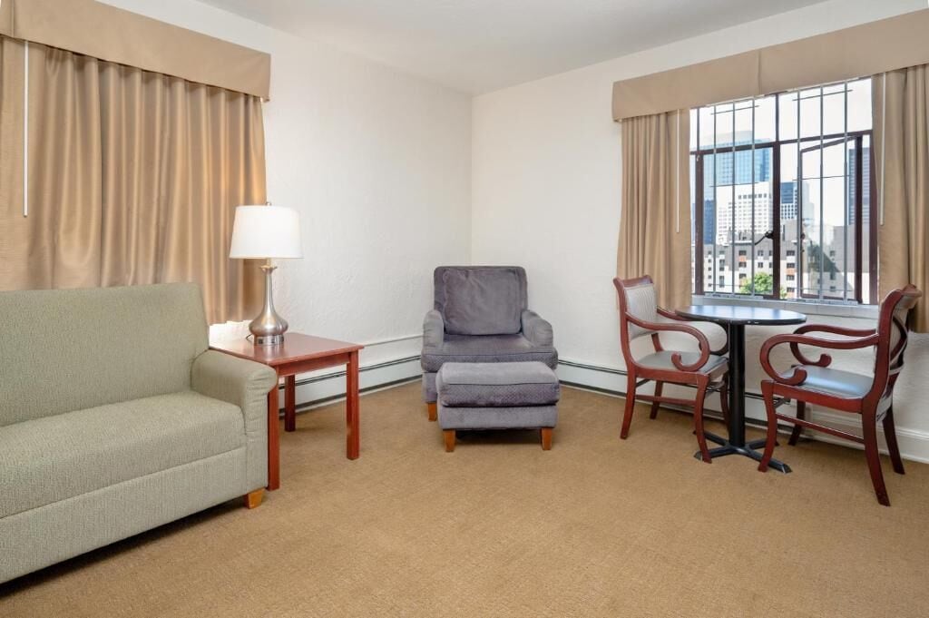 Quarto de hotel com um sofá verde, uma poltrona cinzenta, uma mesinha com abajur e uma janela com vista para edifícios.