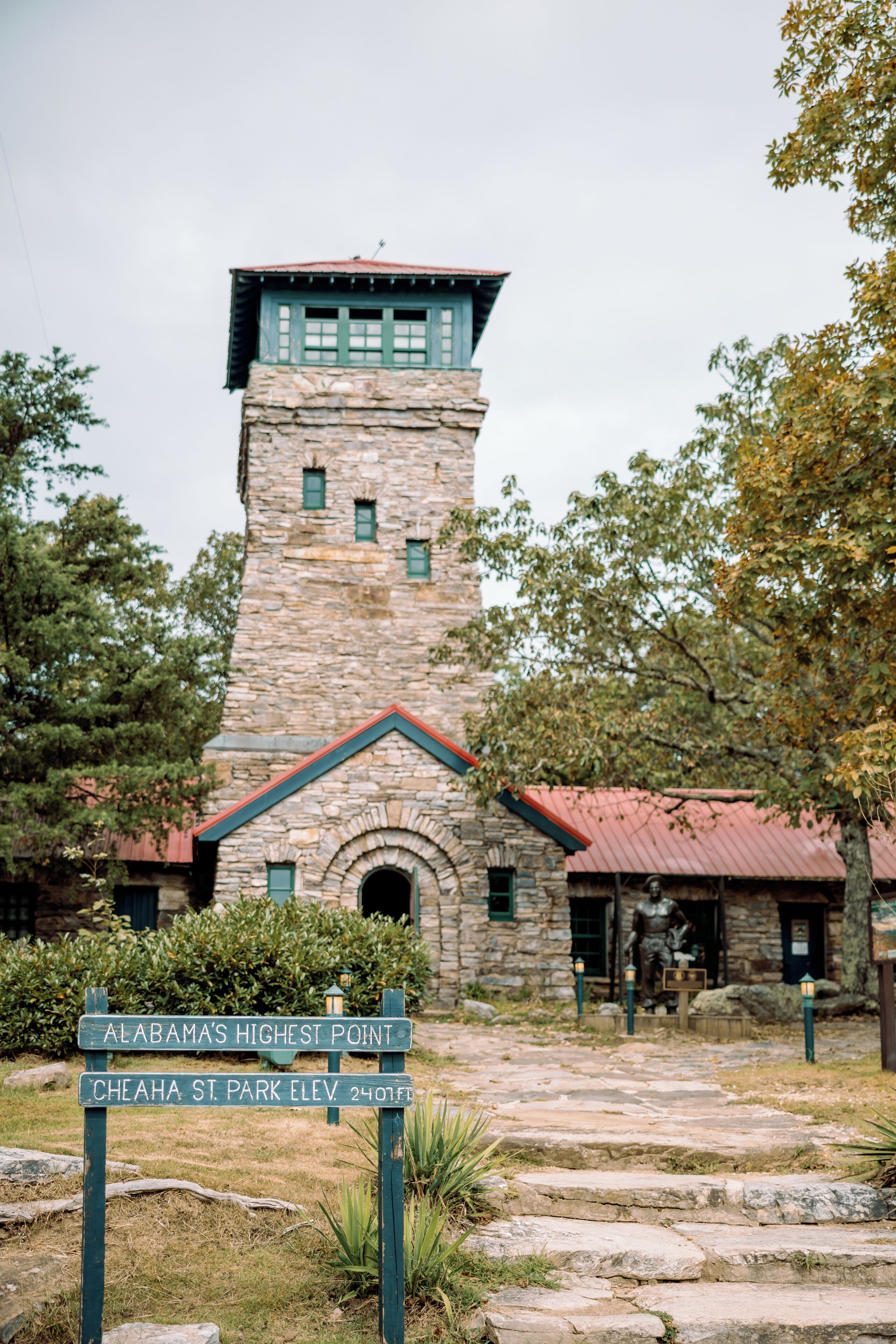 Uma torre de pedra com telhado vermelho, rodeada por árvores e uma placa indicando 'Ponto mais alto do Alabama'.