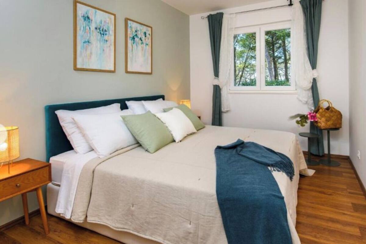 Uma cama bem arrumada com cabeceira azul, mesa de cabeceira com abajur e janela com cortinas.