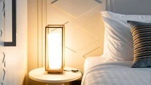 Uma cama bem arrumada com lençóis brancos e almofada listrada, uma mesa de cabeceira redonda com abajur moderno e um design geométrico na parede.