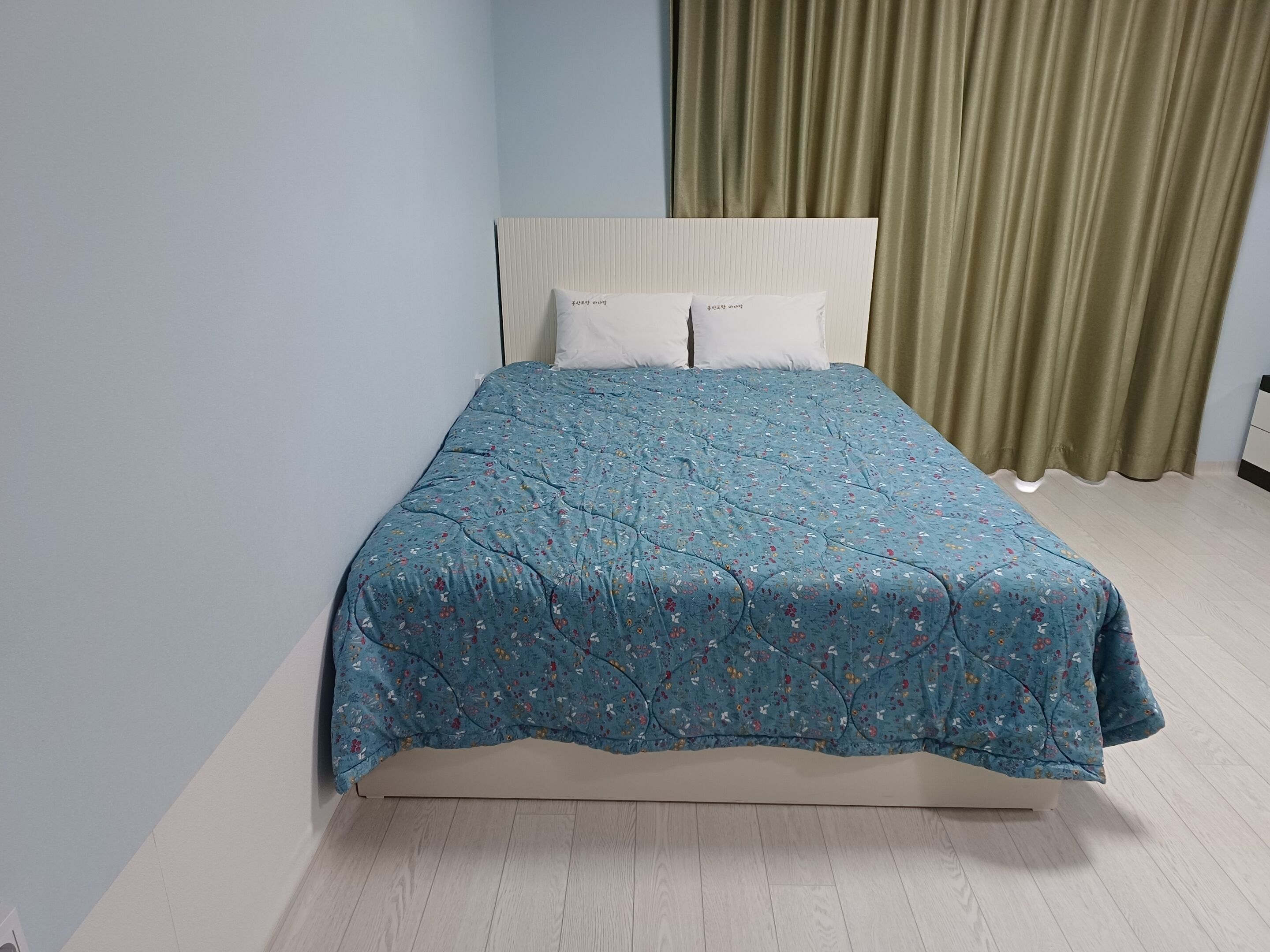 Uma cama com colcha estampada em flor, cabeceira branca e cortina verde.