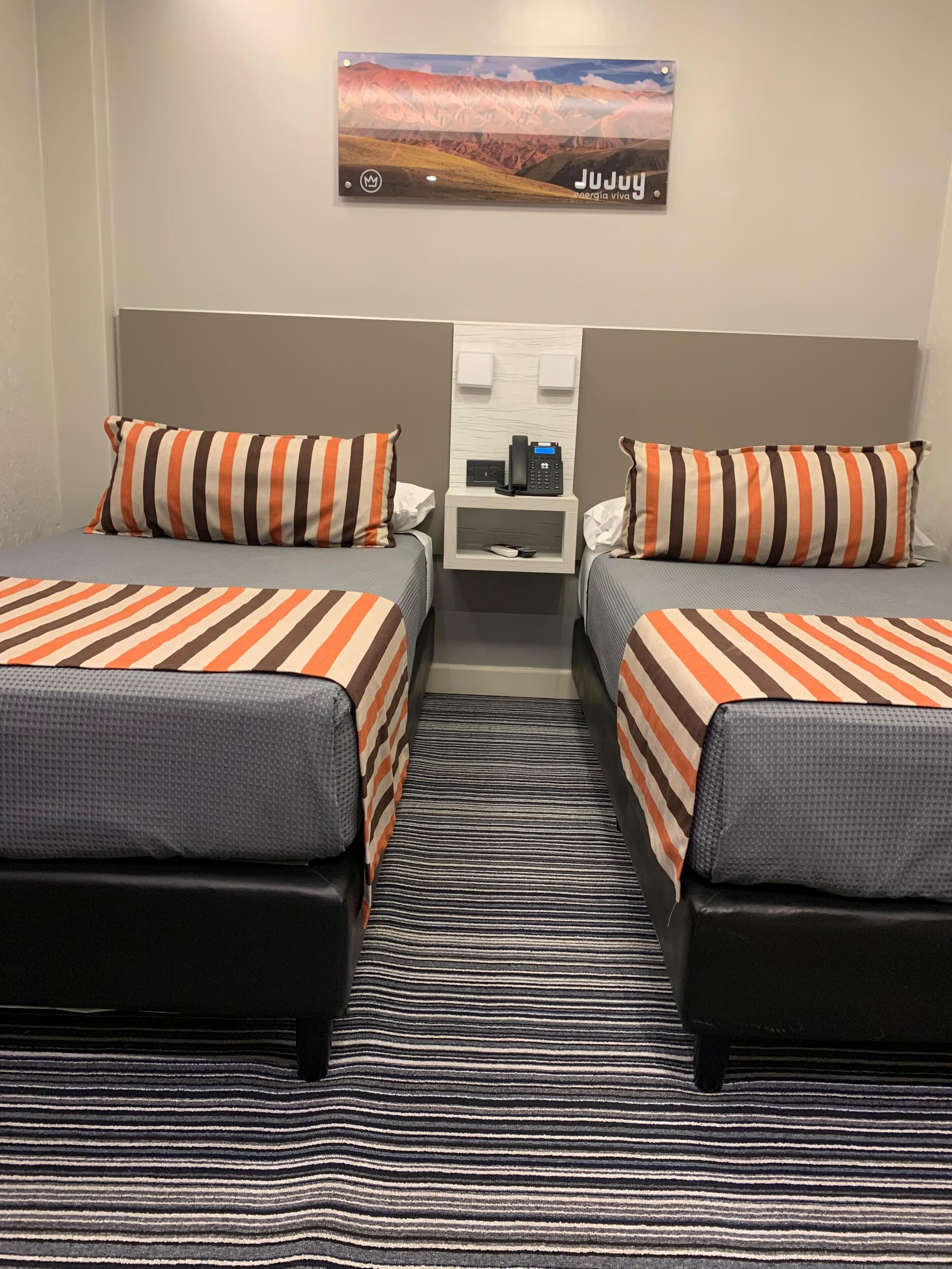Quarto de hotel com duas camas, roupa de cama listrada, tapete estampado, quadro pendurado com paisagem e um telefone em uma mesa de cabeceira.