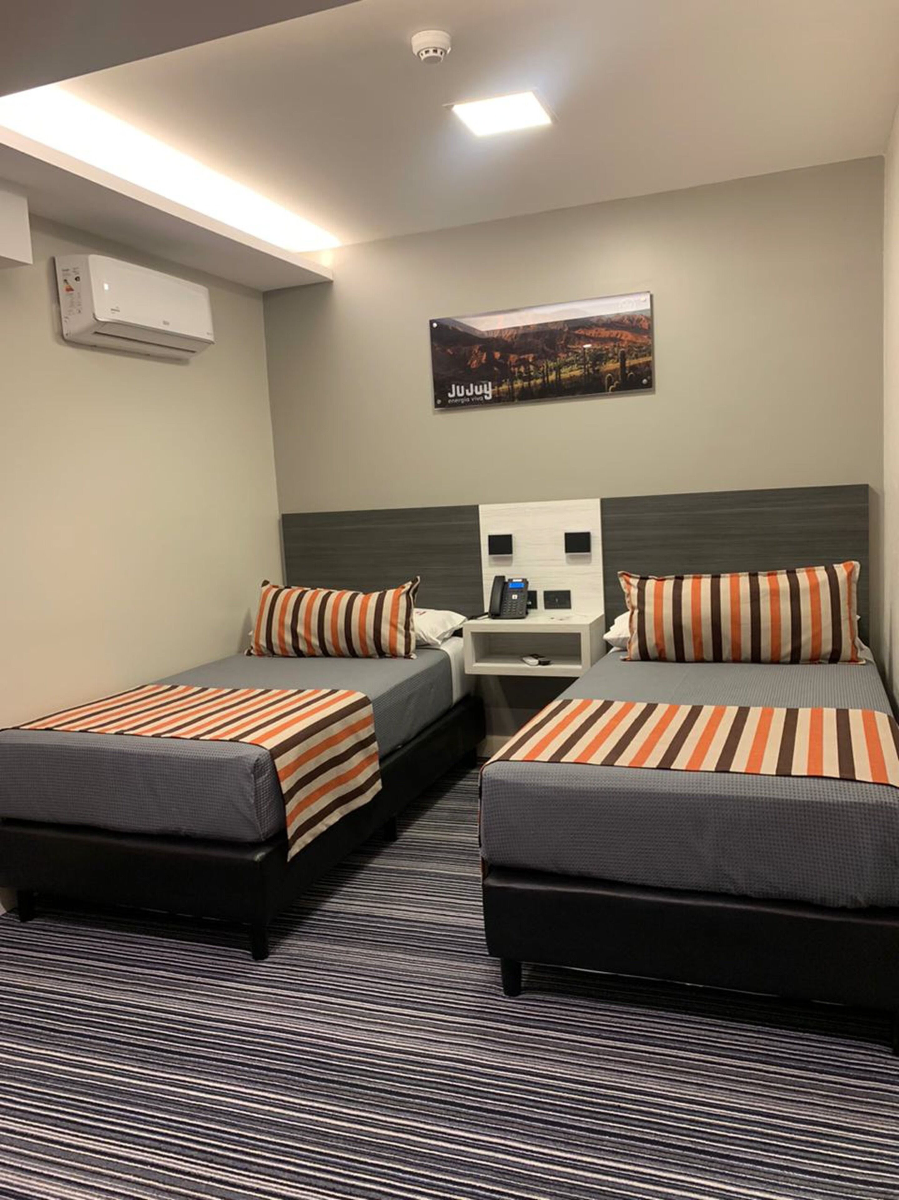 Quarto de hotel com duas camas, uma mesa de cabeceira com telefone, ar condicionado fixado na parede e um quadro emoldurado.