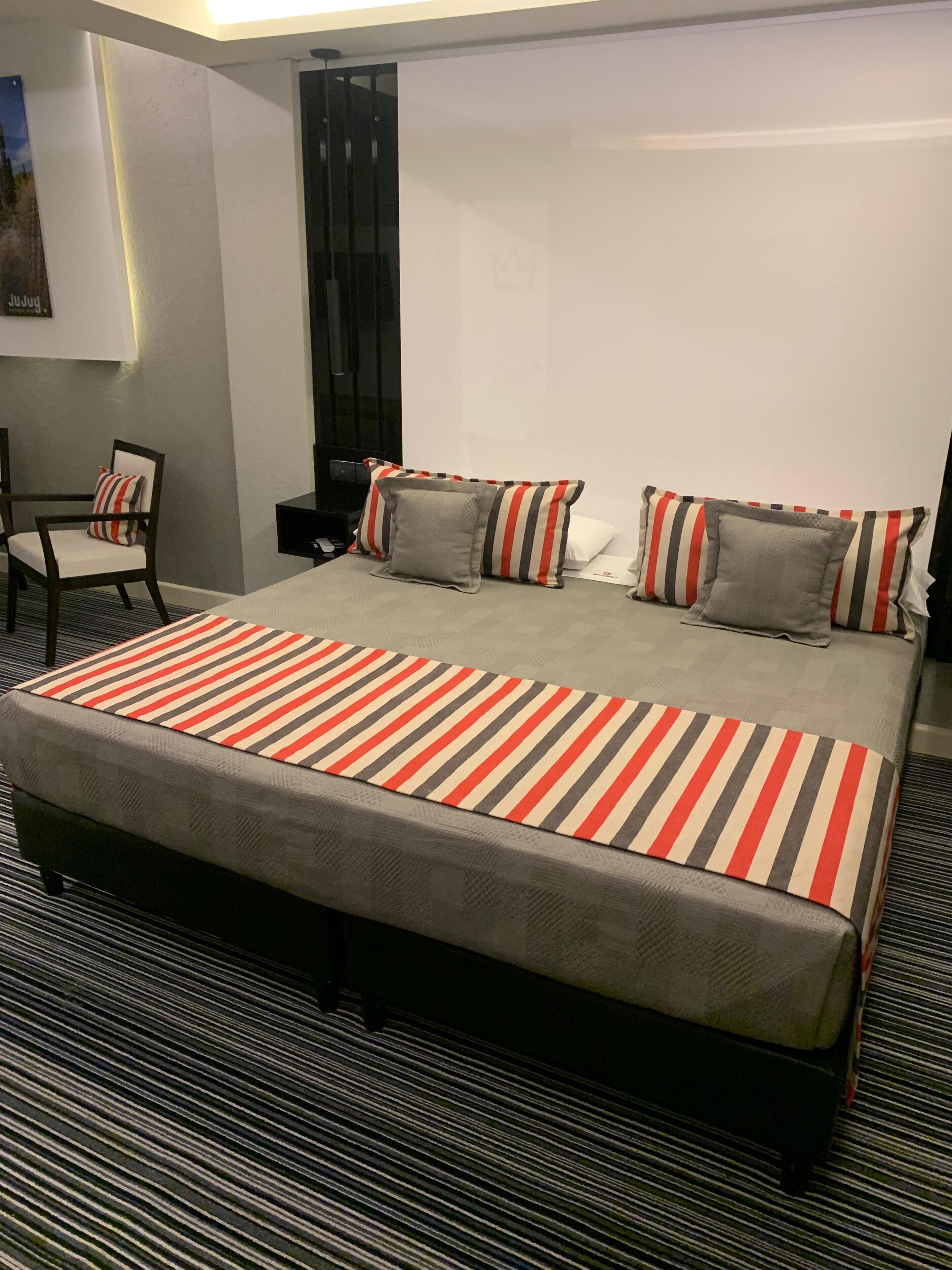 Quarto de hotel com cama grande, roupa de cama listrada, uma cadeira e uma escrivaninha.