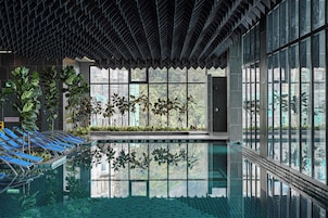 Piscina interna com paredes de vidro transparente, cadeiras de descanso azuis e plantas em vasos.