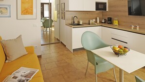 Cozinha moderna com armários brancos, micro-ondas e pia. Há uma área de refeições com mesa e cadeiras, e uma sala de estar com sofá e revista.